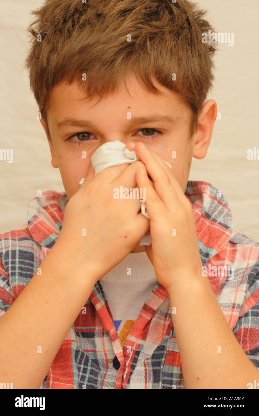 Young boy aged 10 years with cold flu blowing nose with tissue wearing ...