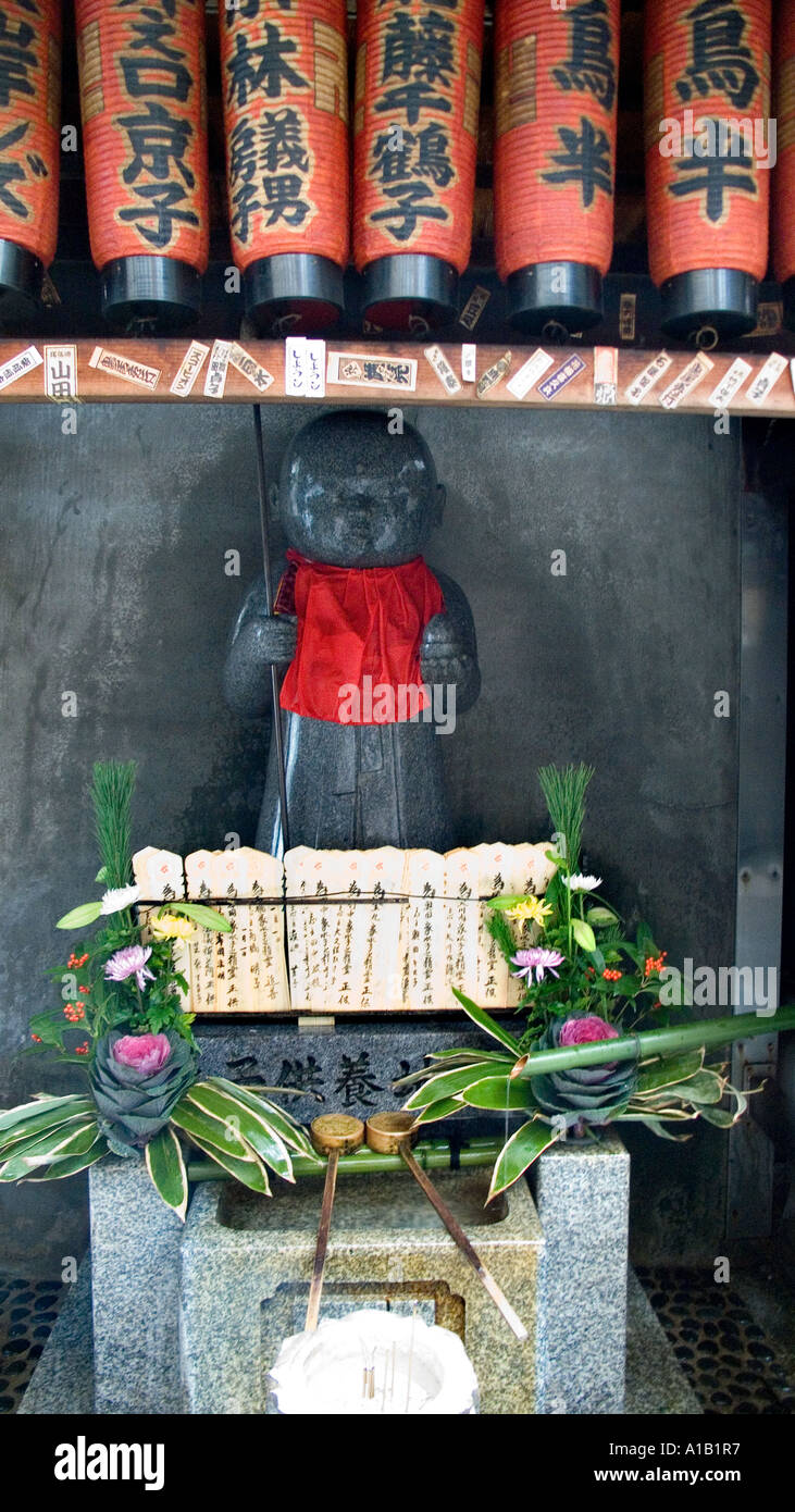 Stone carved Jizo statue in a red bib underneath a row of paper ...