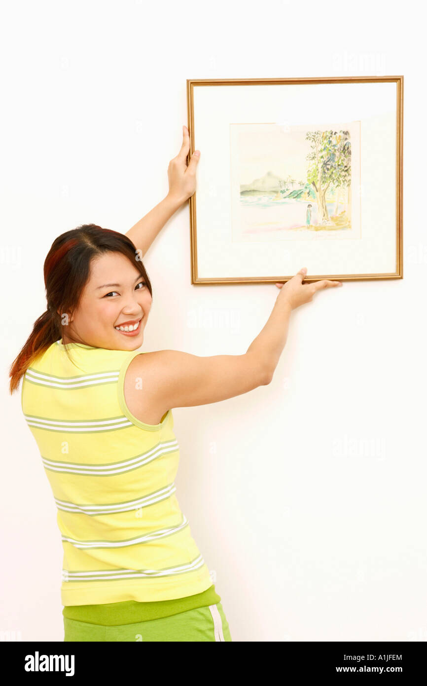 Portrait of a young woman hanging a picture frame on a wall and smiling ...