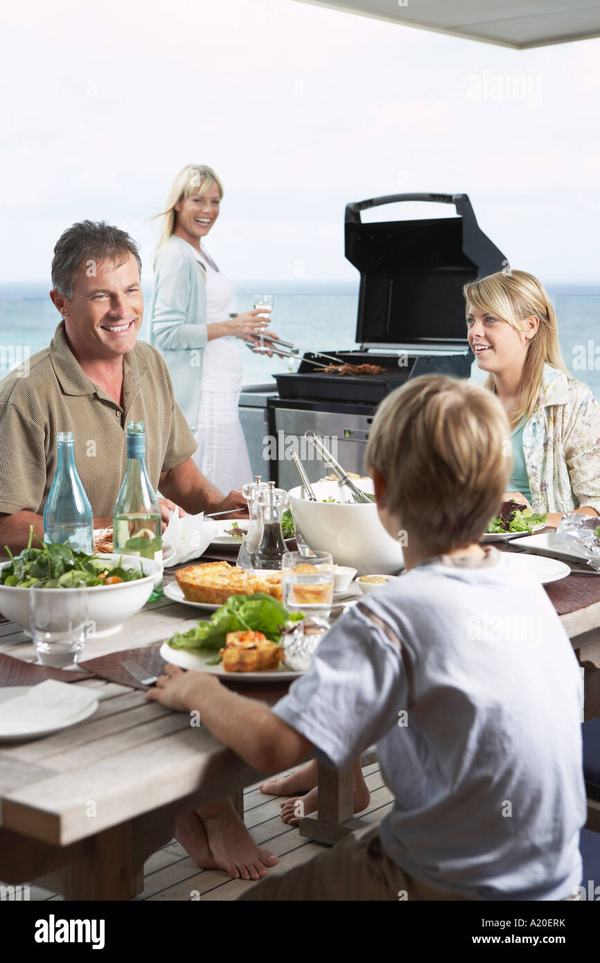 Family sitting around table outside eating Barbecue, mother standing at ...