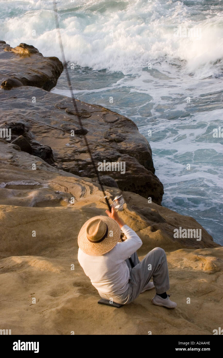A man wearing a straw hat fishes along a rocky shoreline, with waves ...