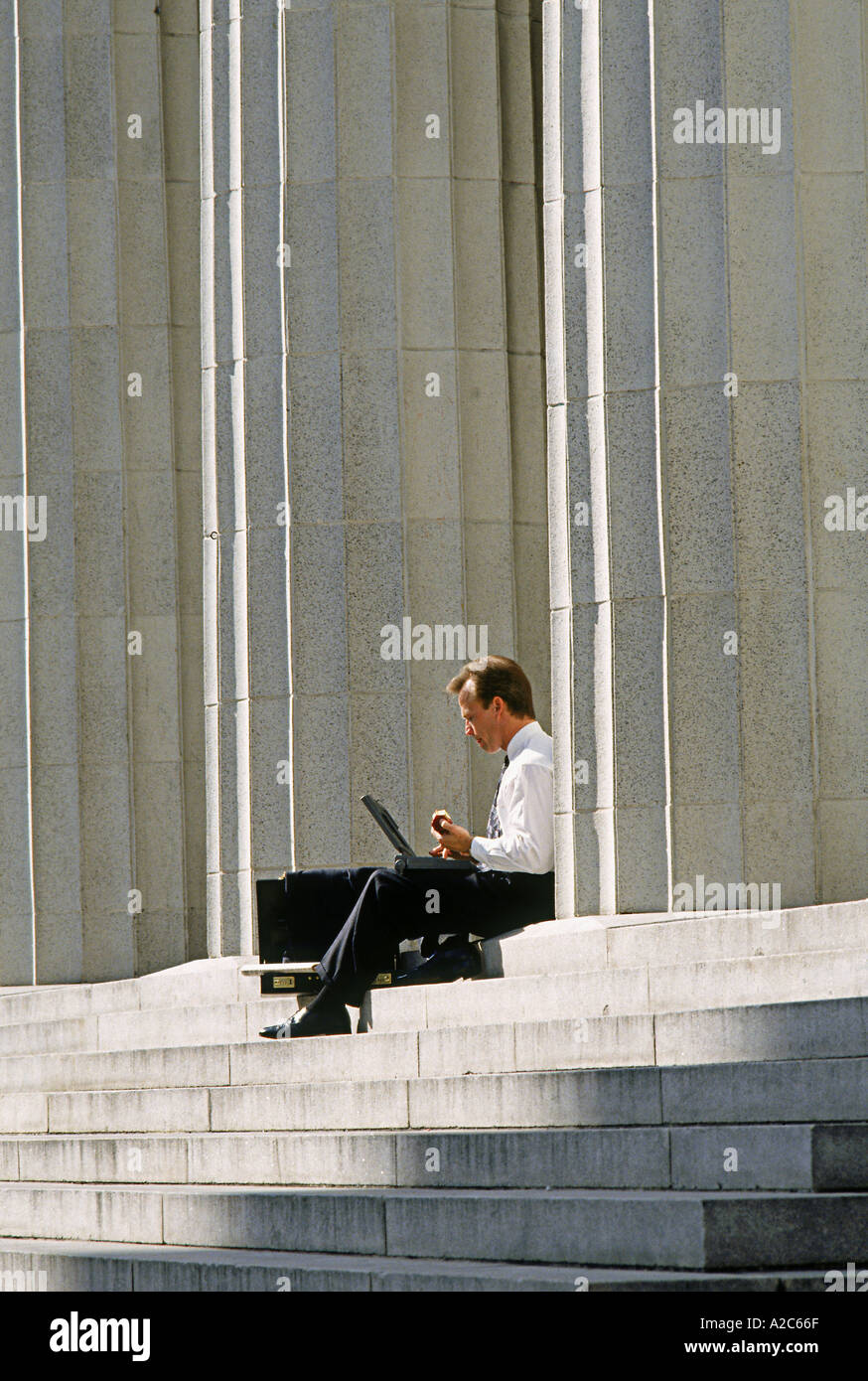 A businessman using a lap top computer is sitting on the steps of a ...