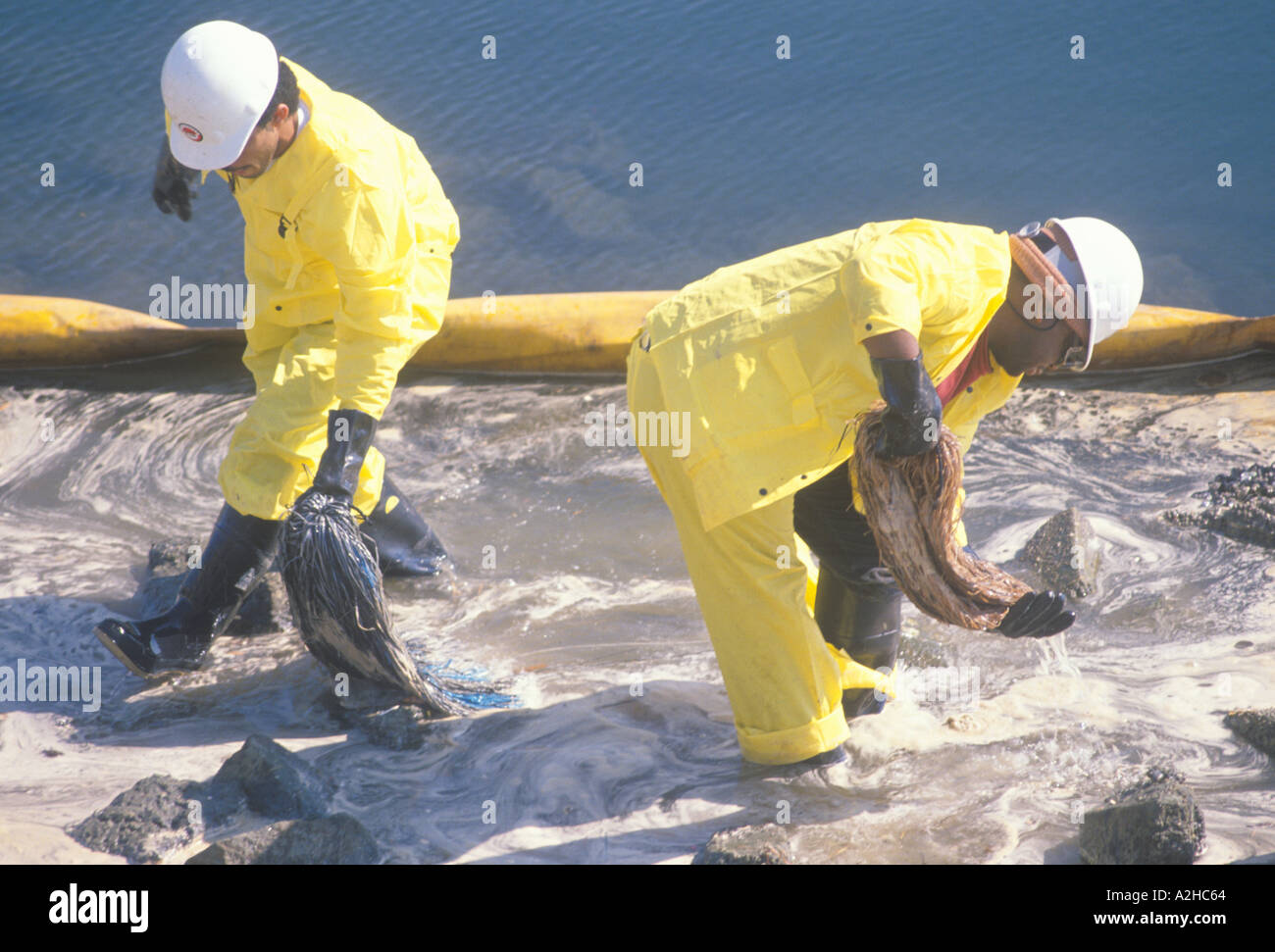 Two oil cleanup workers wade in oily water between a yellow oil barrier ...
