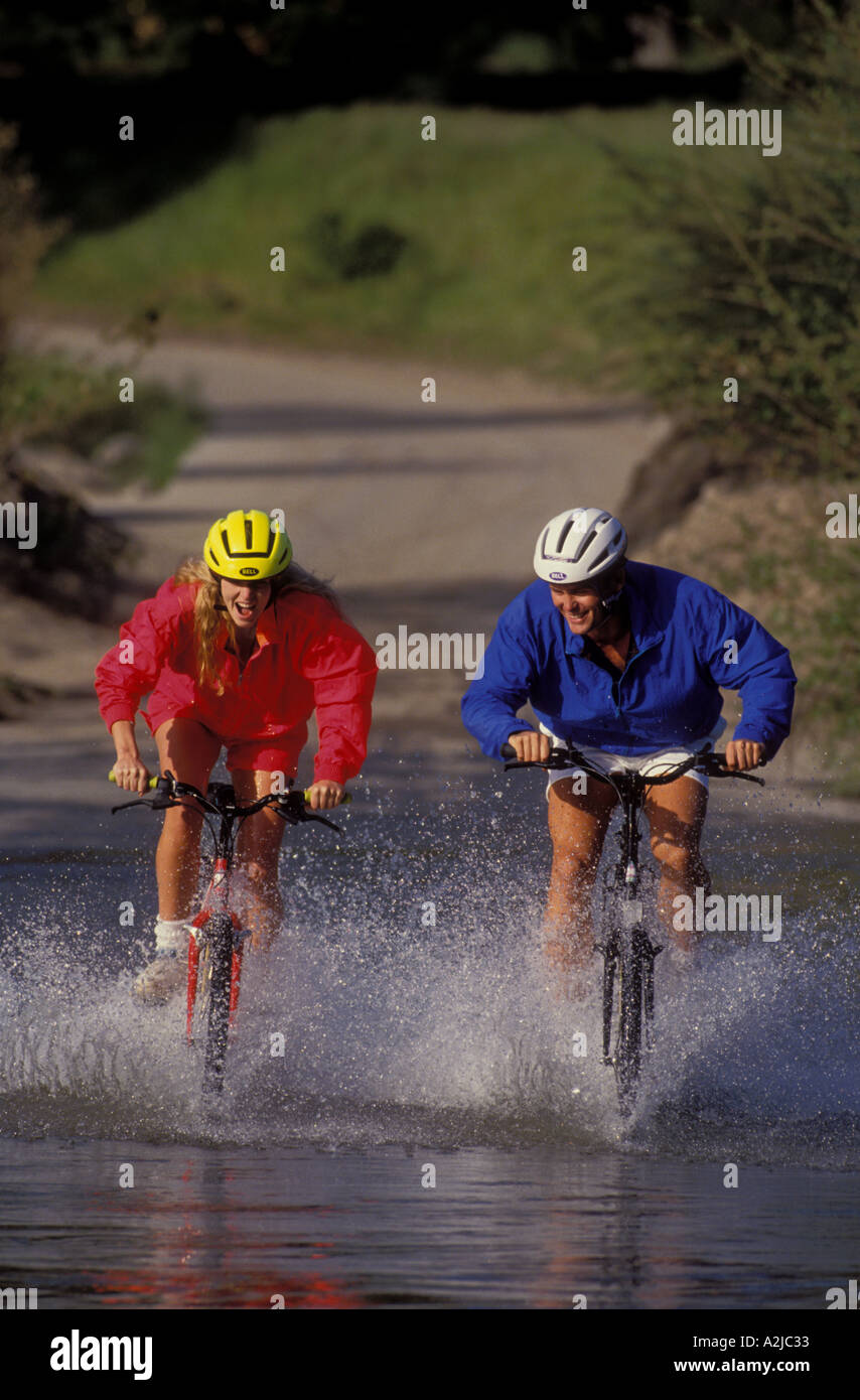 Man and woman riding bikes side by side splash through water on a cross ...
