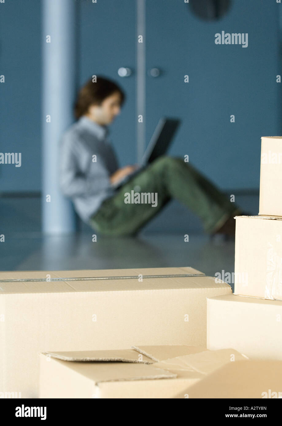 Young man sitting on floor using laptop, cardboard boxes in foreground ...