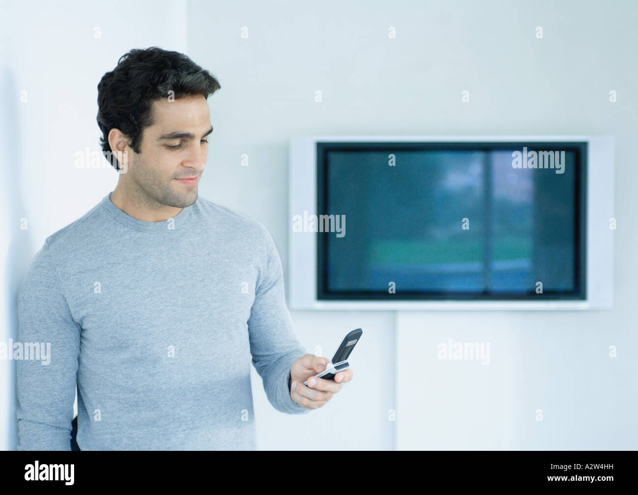 Man standing, looking at cell phone, widescreen TV on wall in ...