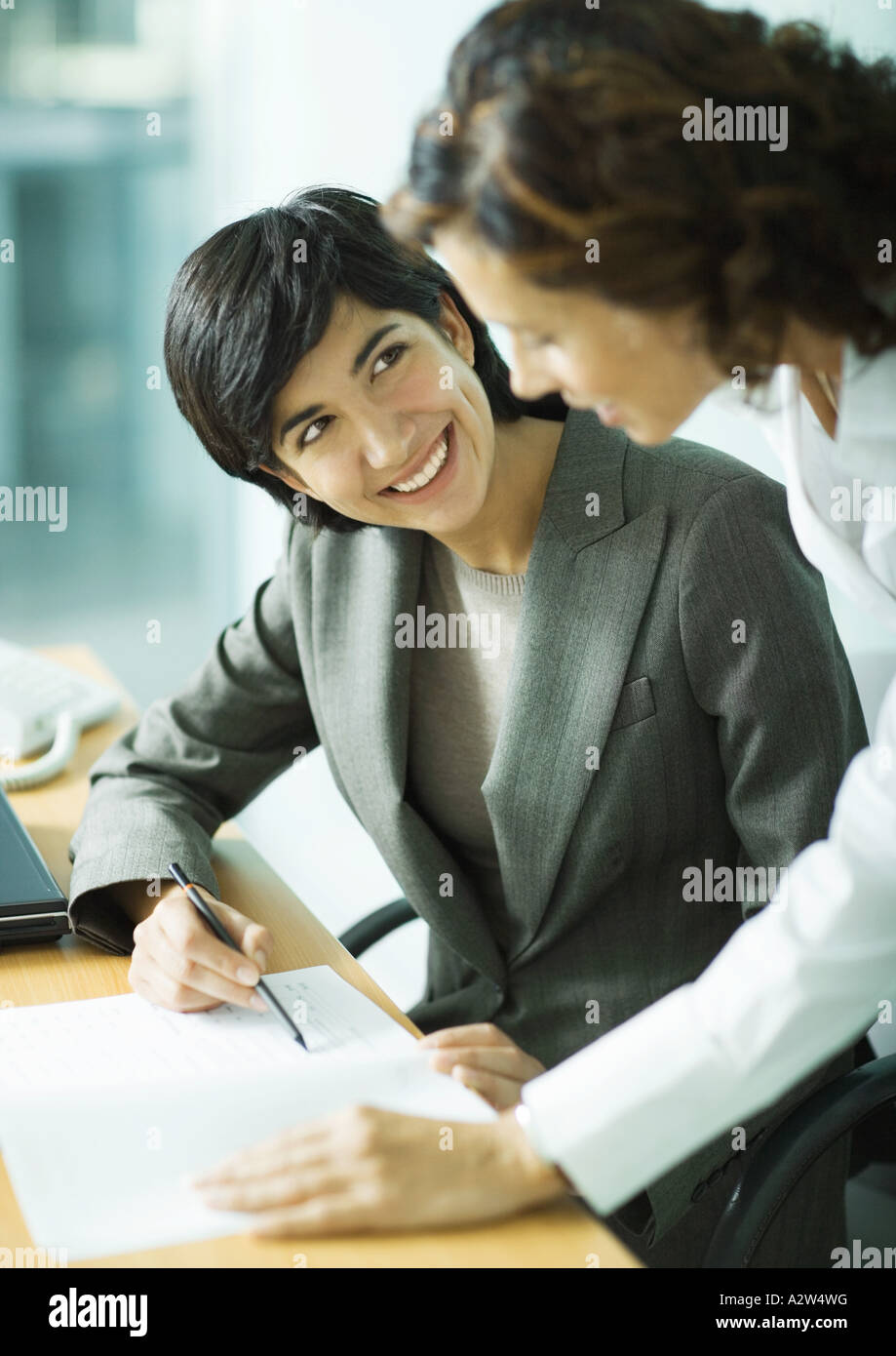 Woman sitting as desk, pointing to document and smiling at second woman ...