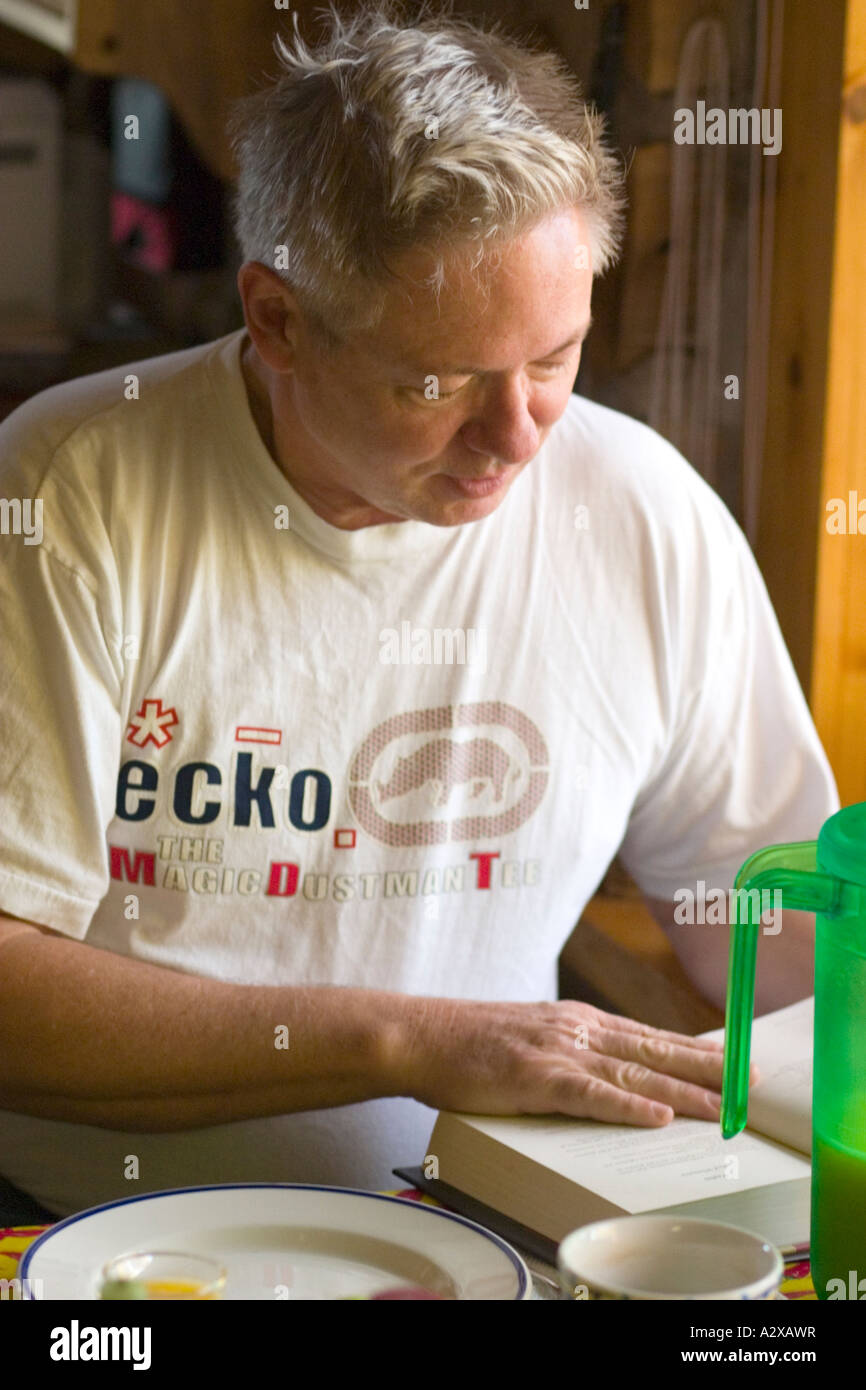 Man age 59 in solitude reading book at breakfast in his cabin. Cable ...