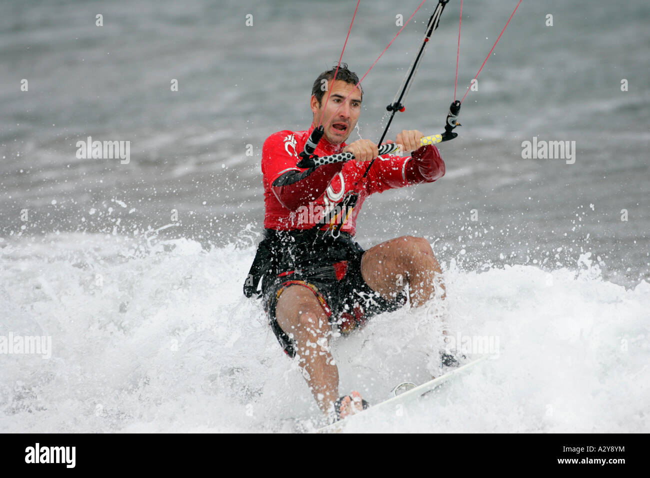 male kitesurfer in red lifts his board through a wave in the water off ...