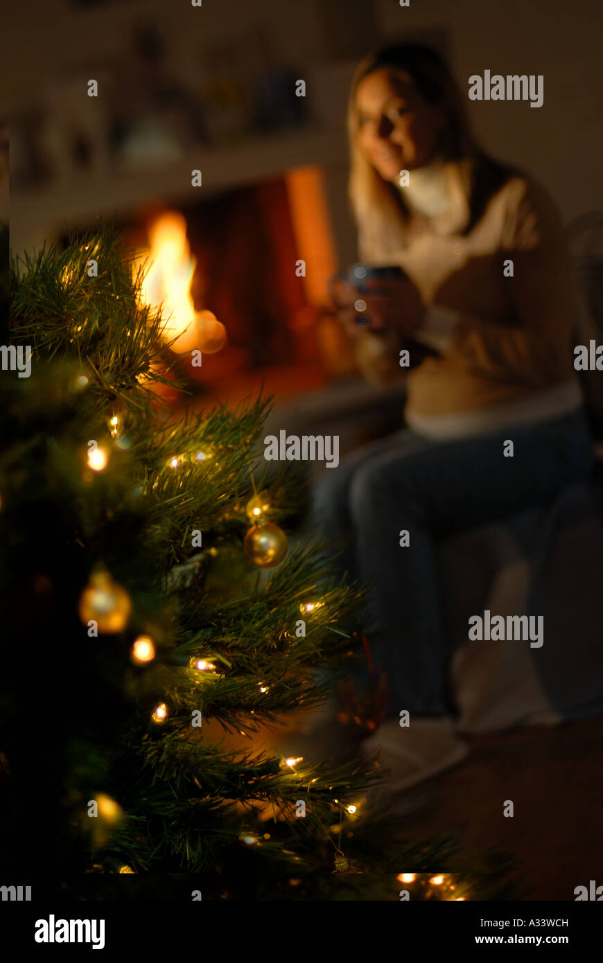 Young woman sat in front of open fire at christmas tree with hot drink ...