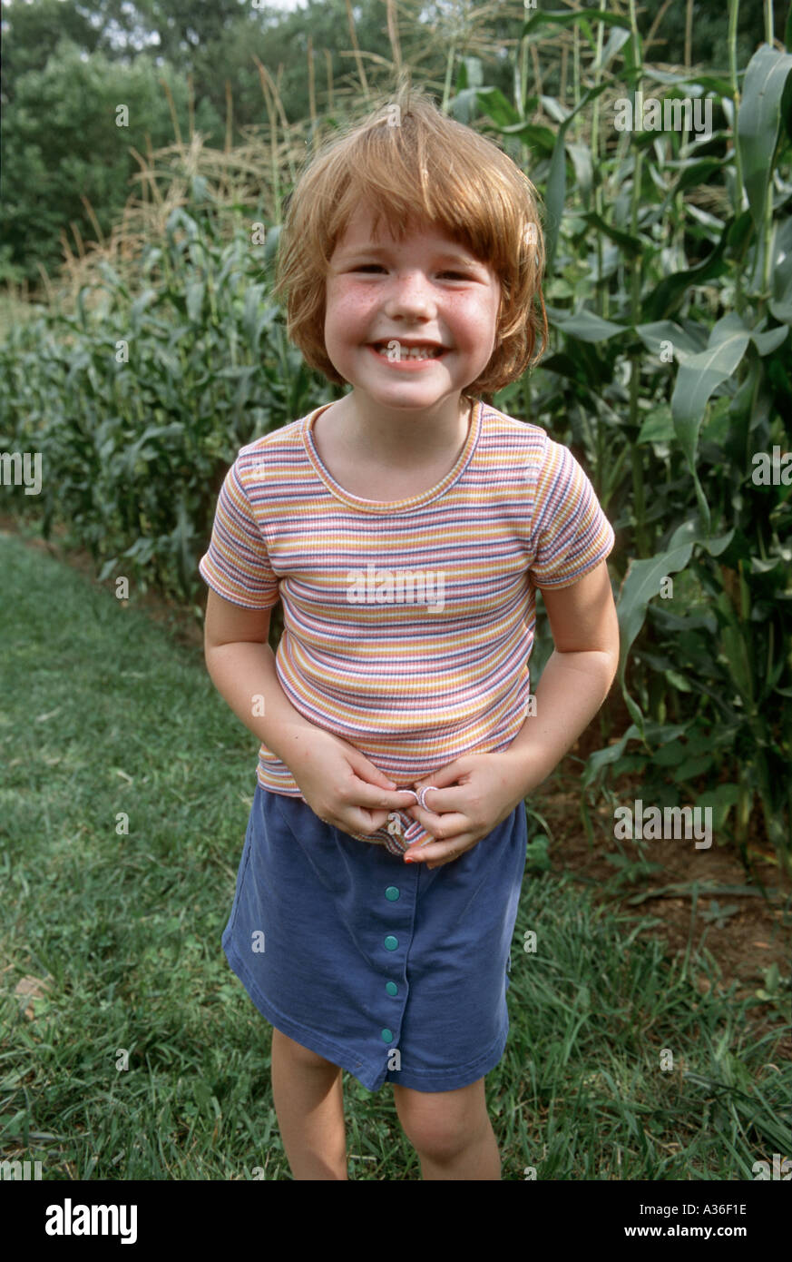 A cute young girl with rosy cheeks in standing by a field of corn an is ...