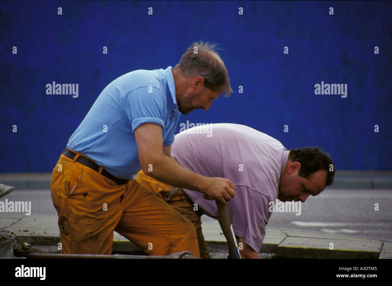 Two men in coloured shirts and orange trousers digging a hole in the ...