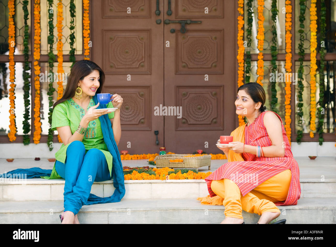 Two young women sitting at doorstep of a house and holding cups of tea ...