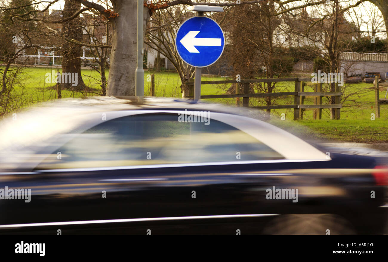 a bright blue turn left road sign with a car speeding past trees in the ...