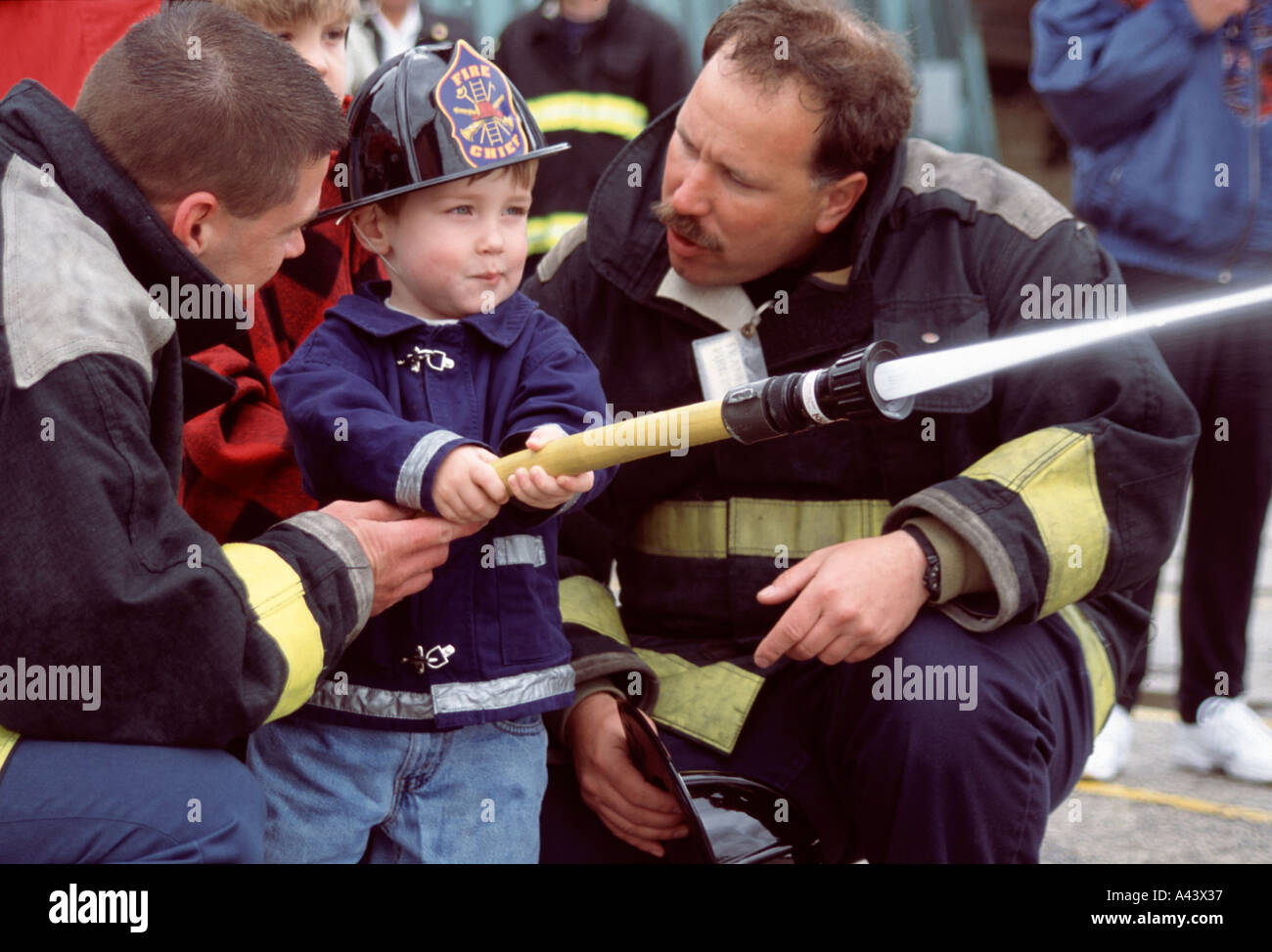 Two fire fighters helping young child with hose to put out fire ...
