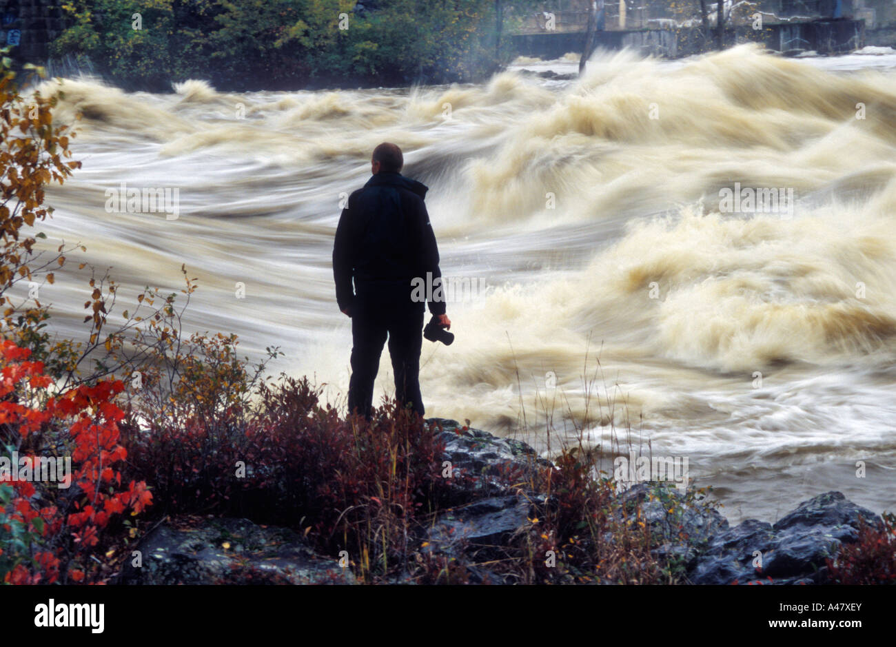 A photographer silhouetted against a river in full flood after autumnal ...