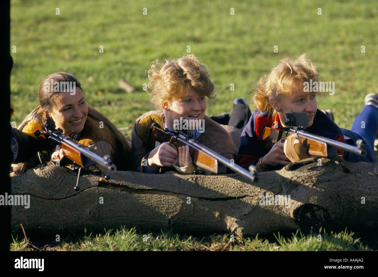 3 smiling young women lie on grass and rest their guns on a thick tree ...