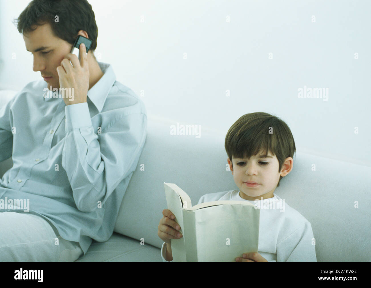 Man sitting on sofa talking on cell phone, boy sitting next to him ...