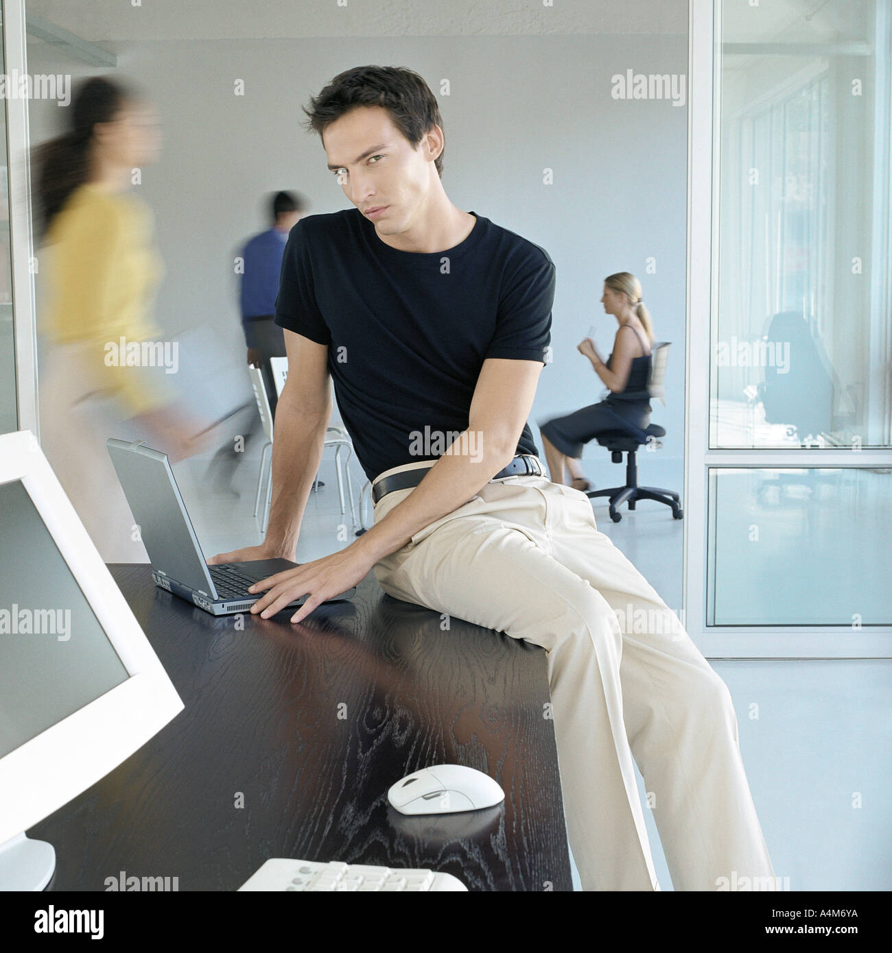 Man sitting on edge of desk in busy office Stock Photo - Alamy