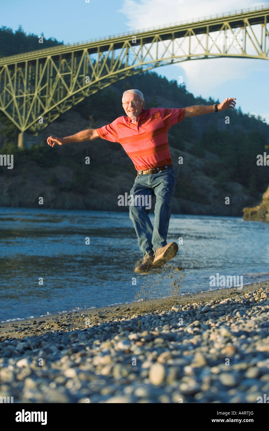 Senior man clicking heels in midair on beach Deception Pass Bridge ...