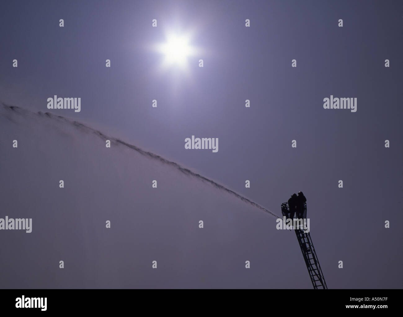 Two fireman on a high lift platform testing a hose against a bright sun ...