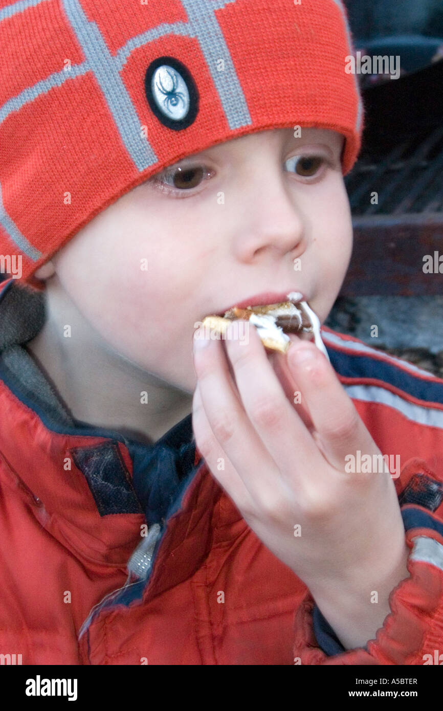 Boy age 6 eating a smore by squishing heated marshmallow and chocolate ...