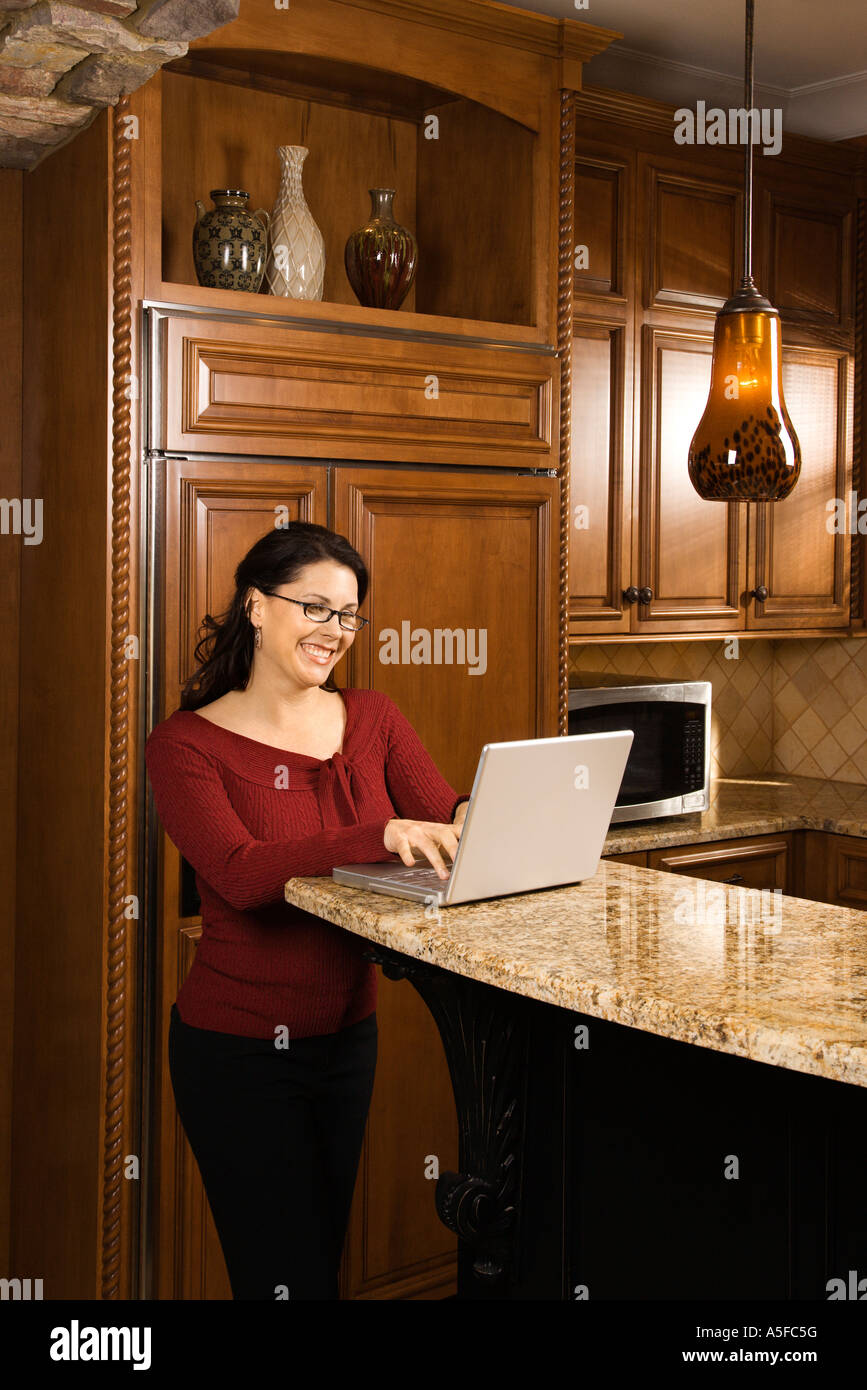 Caucasian woman standing at kitchen counter typing on laptop computer ...