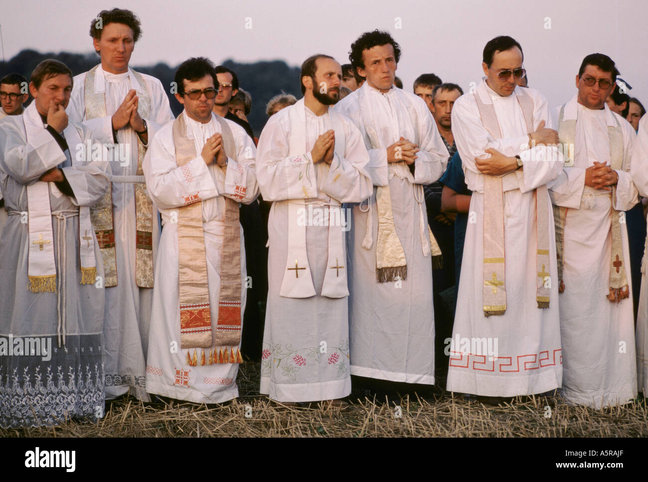 LINE OF SEVEN PRIESTS STAND PRAYING AMONGST POLISH PILGRIMS AT EVENING ...