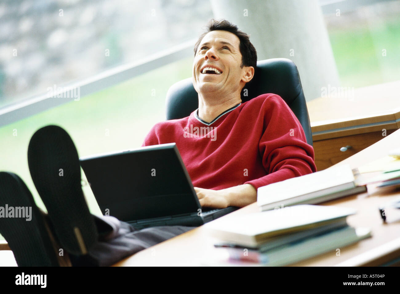 Man sitting at desk with feet up, holding laptop on lap, laughing Stock ...