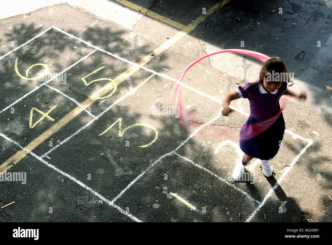 While playing hopscotch, a young girl twirls her hula hoop in a ...