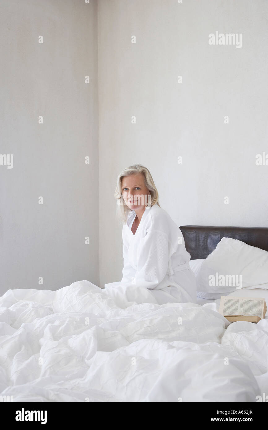 Mature woman in bathrobe, sitting up in bed, smiling, next to open book ...