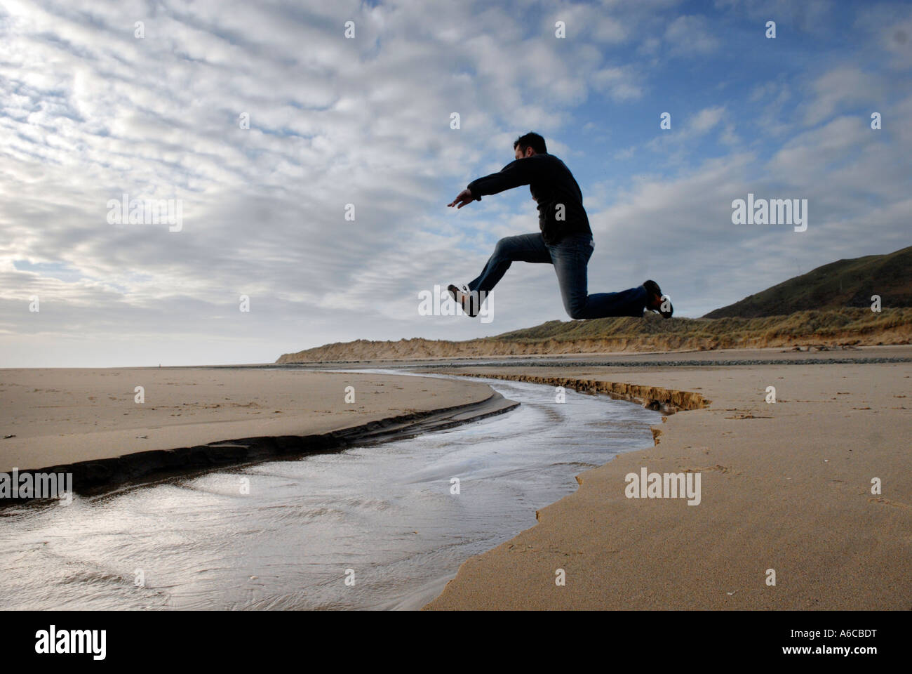 A man jumping over a river of sea water on Aberdovey beach Wales Stock ...