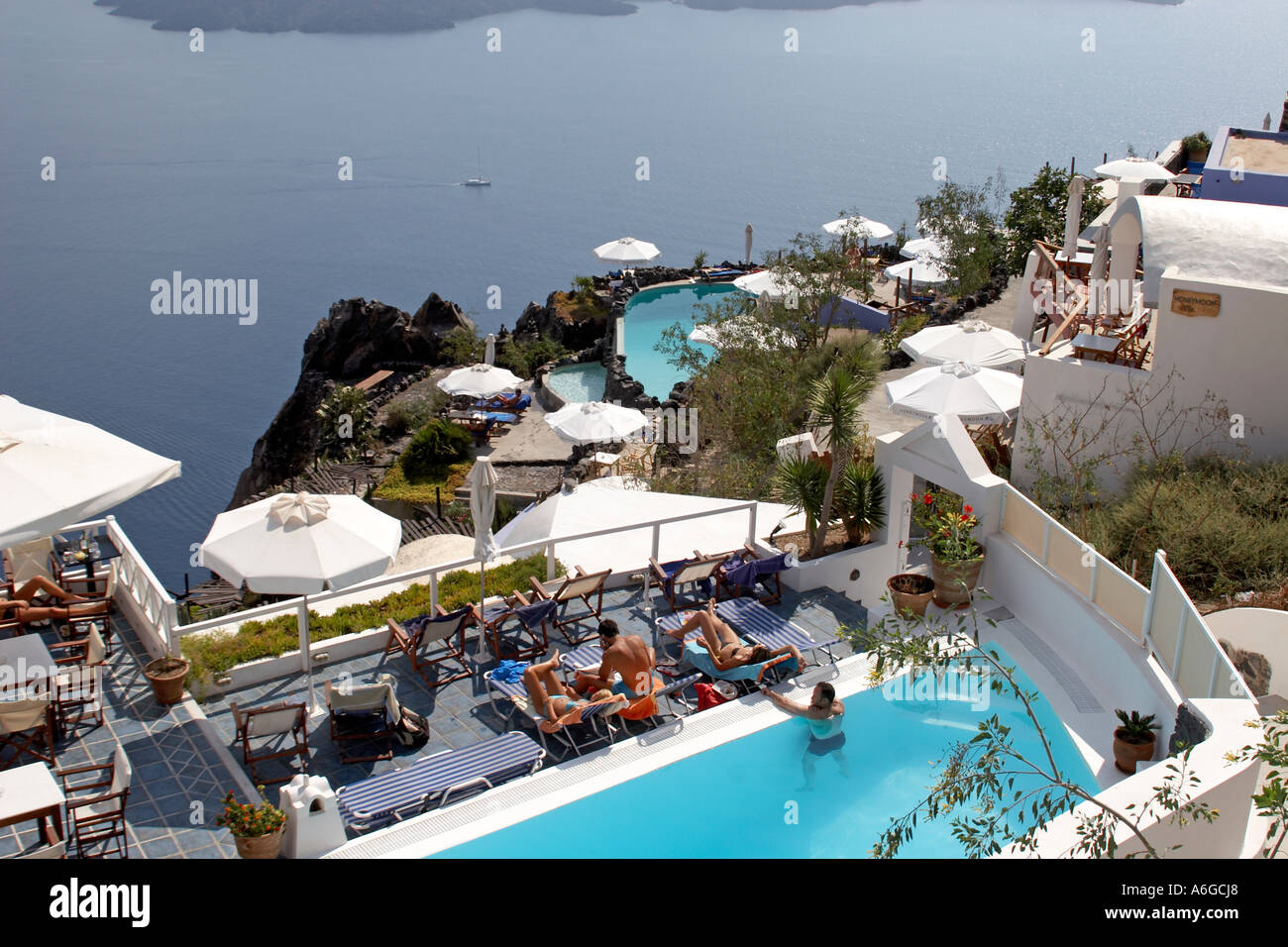 Hotel pool with people sunbathing and the Aegean sea beyond Imerovigli ...