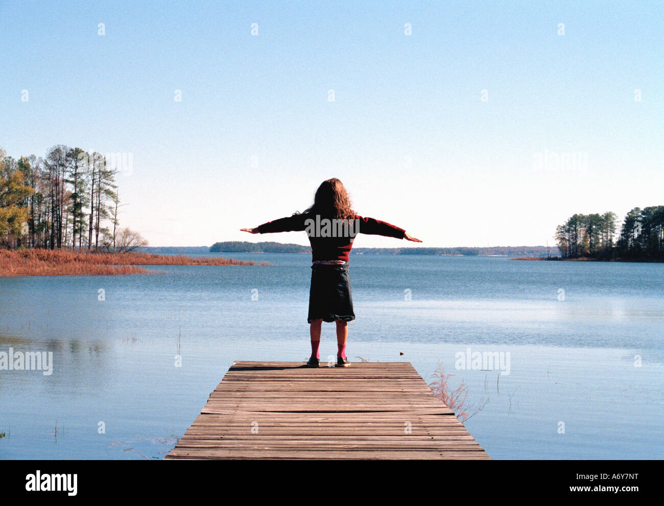 Young girl standing at the edge of a wooden jetty with her arms ...