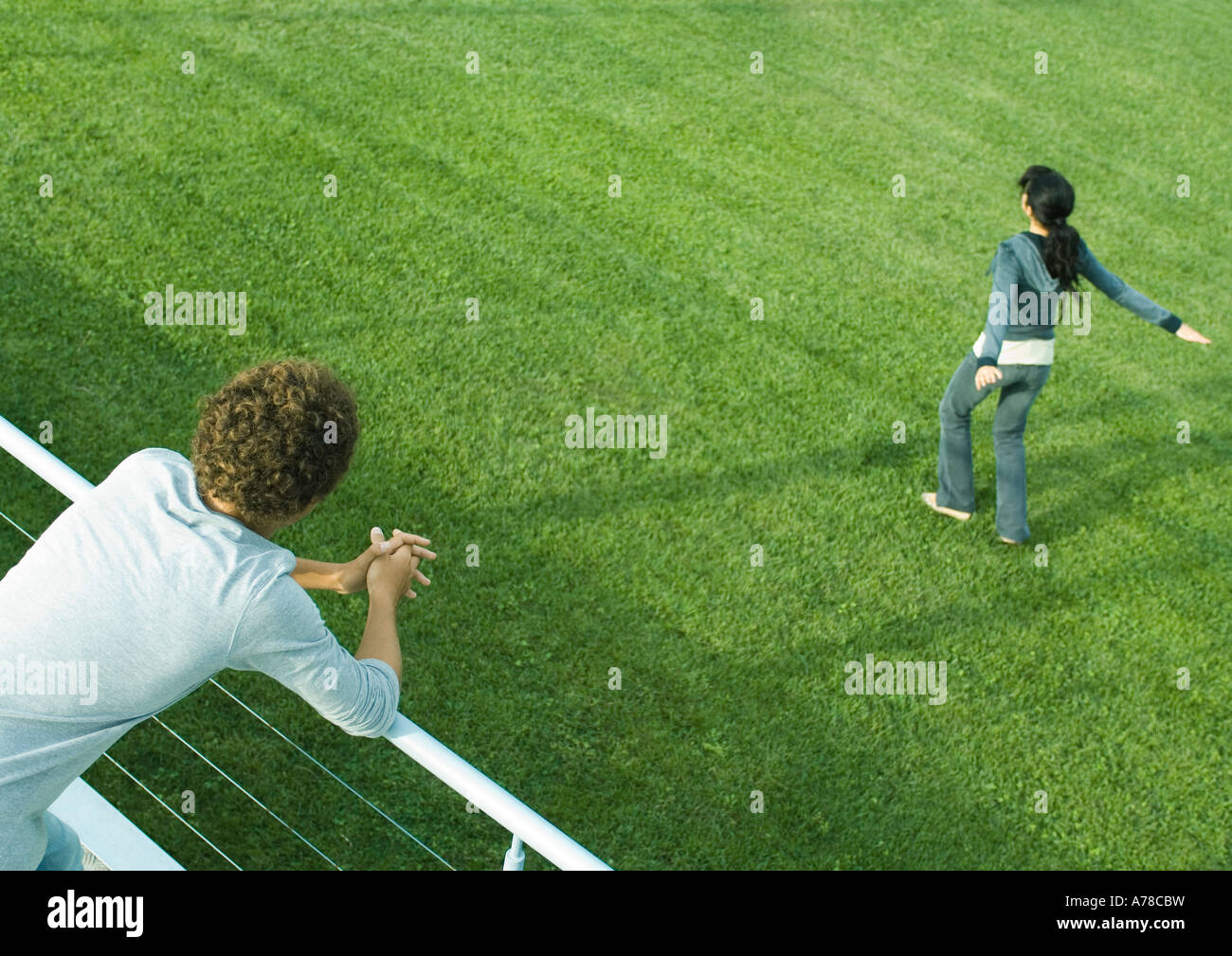 Teenage boy leaning on railing, watching teenage girl spinning on grass ...