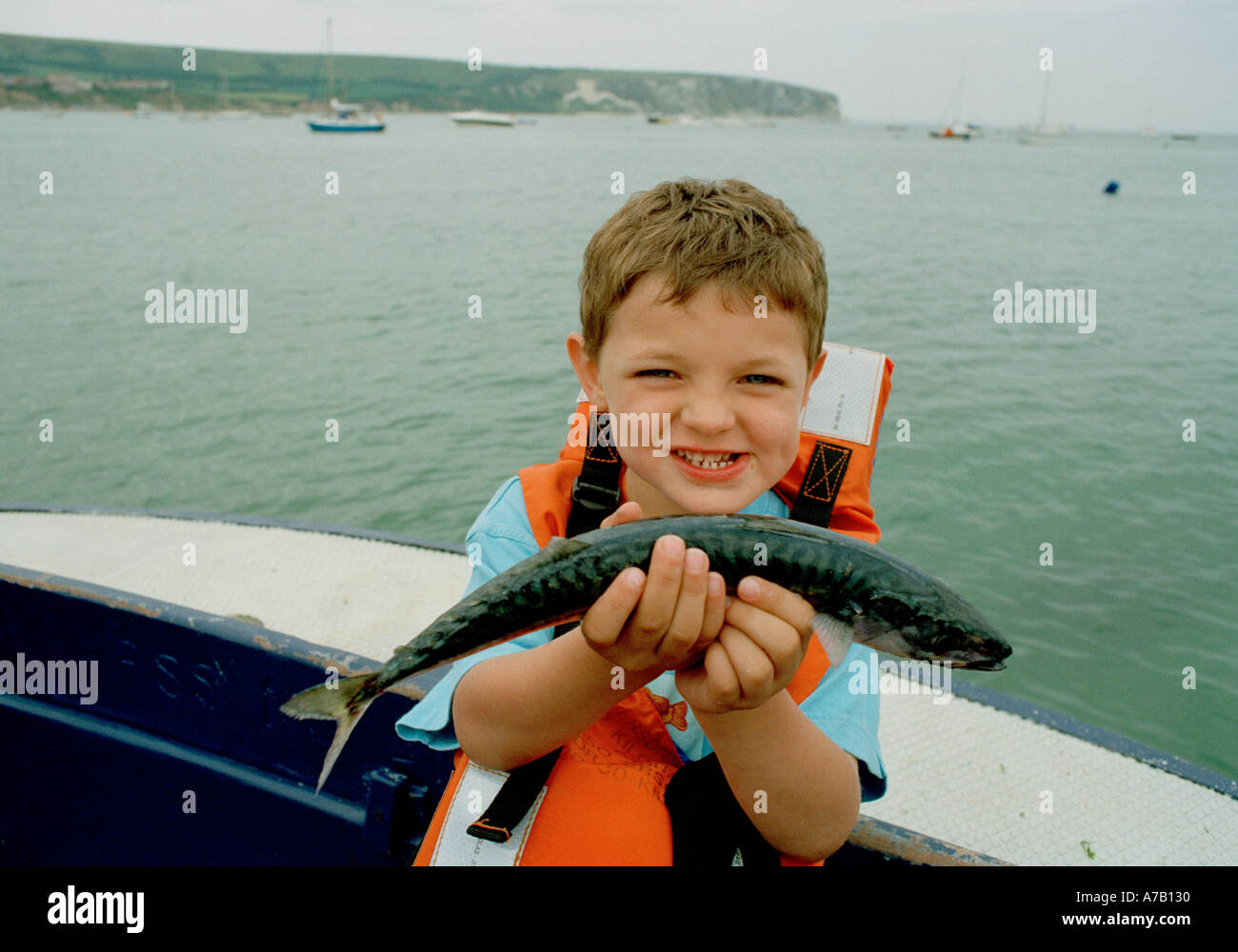 Small boy on boat holding a fish to camera with the sea and headland in ...
