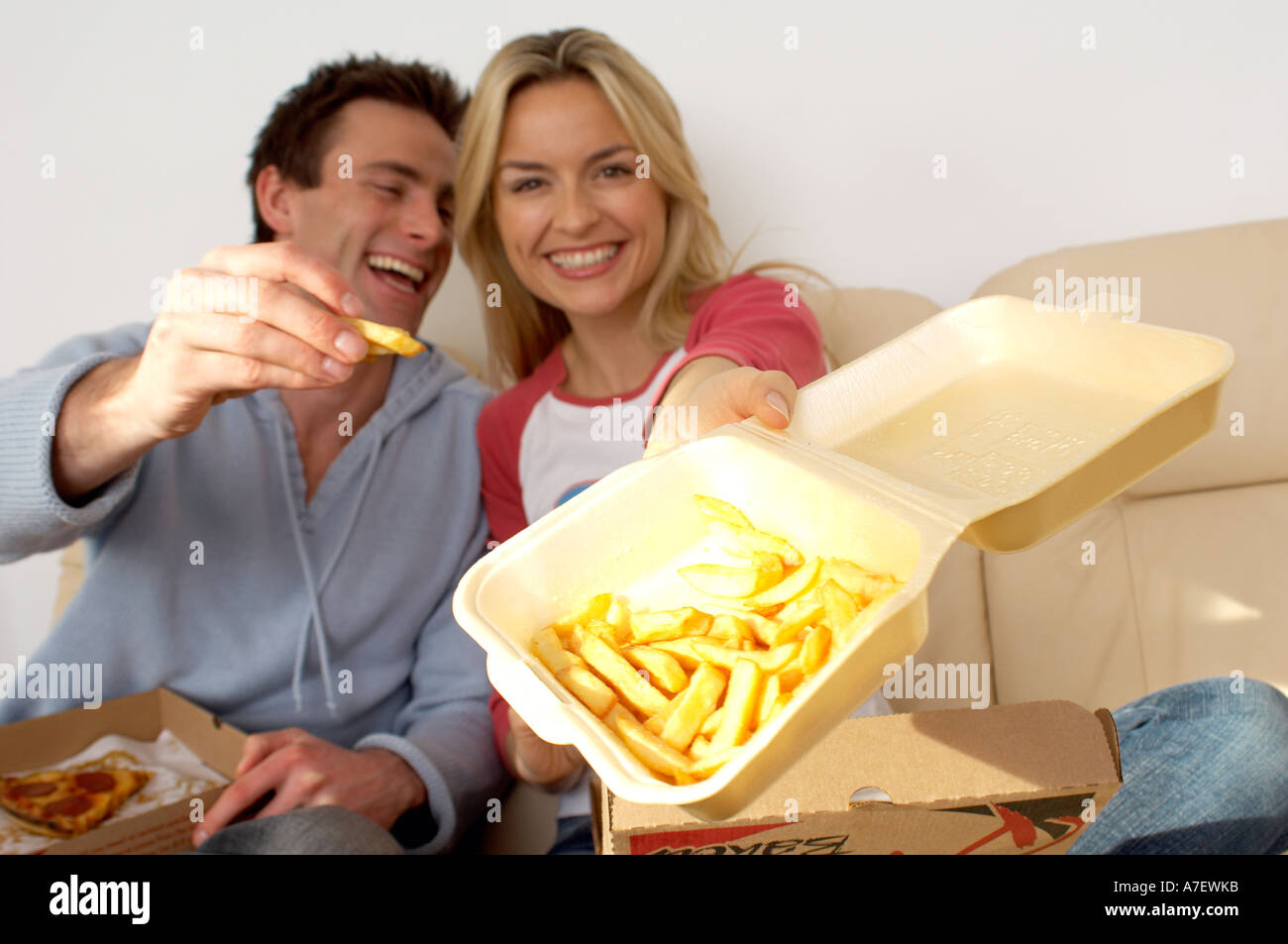 couple sitting on sofa eating pizza and chips offering chips towards ...