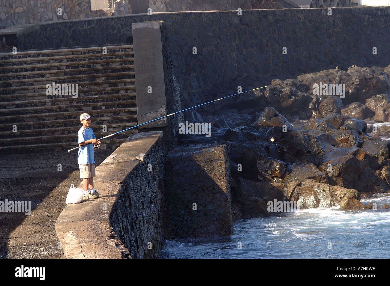 Boy standing on harbour wall in cap reeling in fishing rod trying to ...