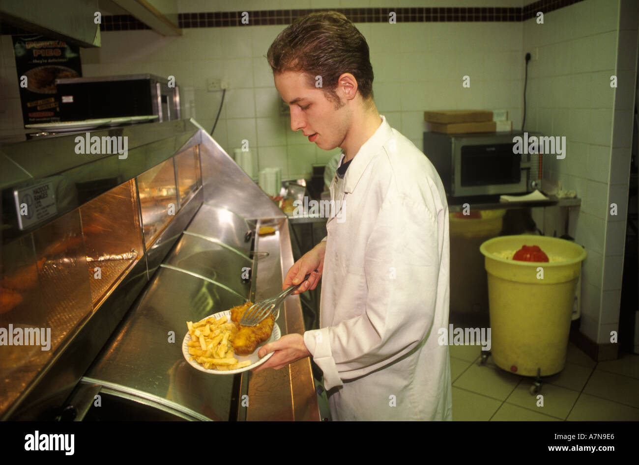 Fish and Chip shop young man putting piece of cod on a plate along with ...
