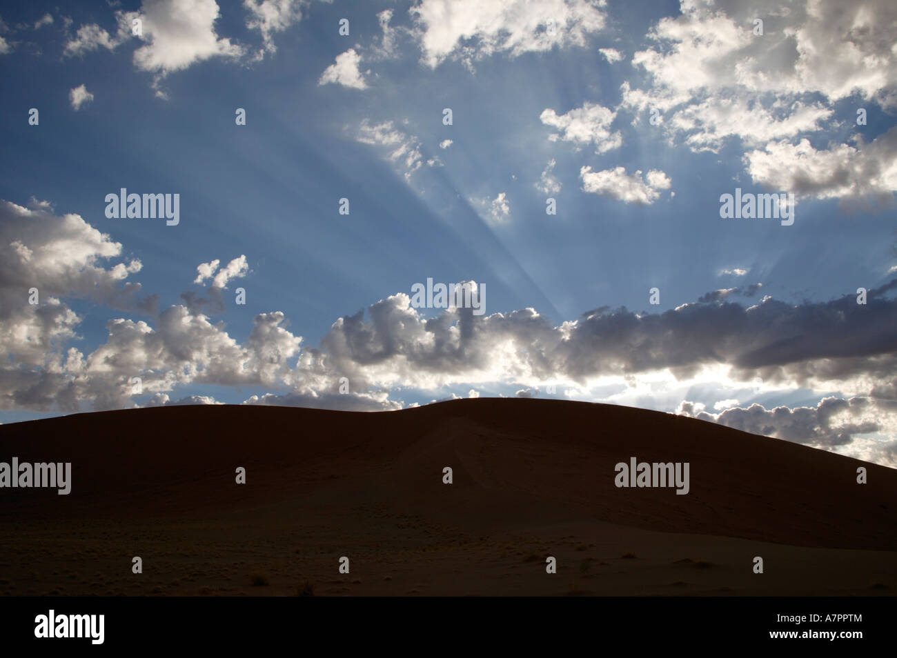 Silhouetted sand dune against a dramatic sky with the sun rays shining ...