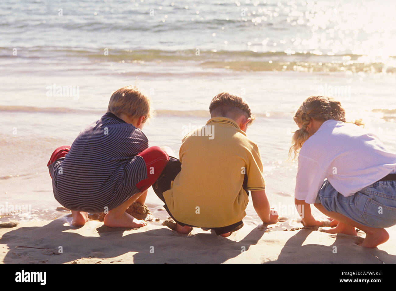 portrait of three children squatting in the sand collecting mussels ...