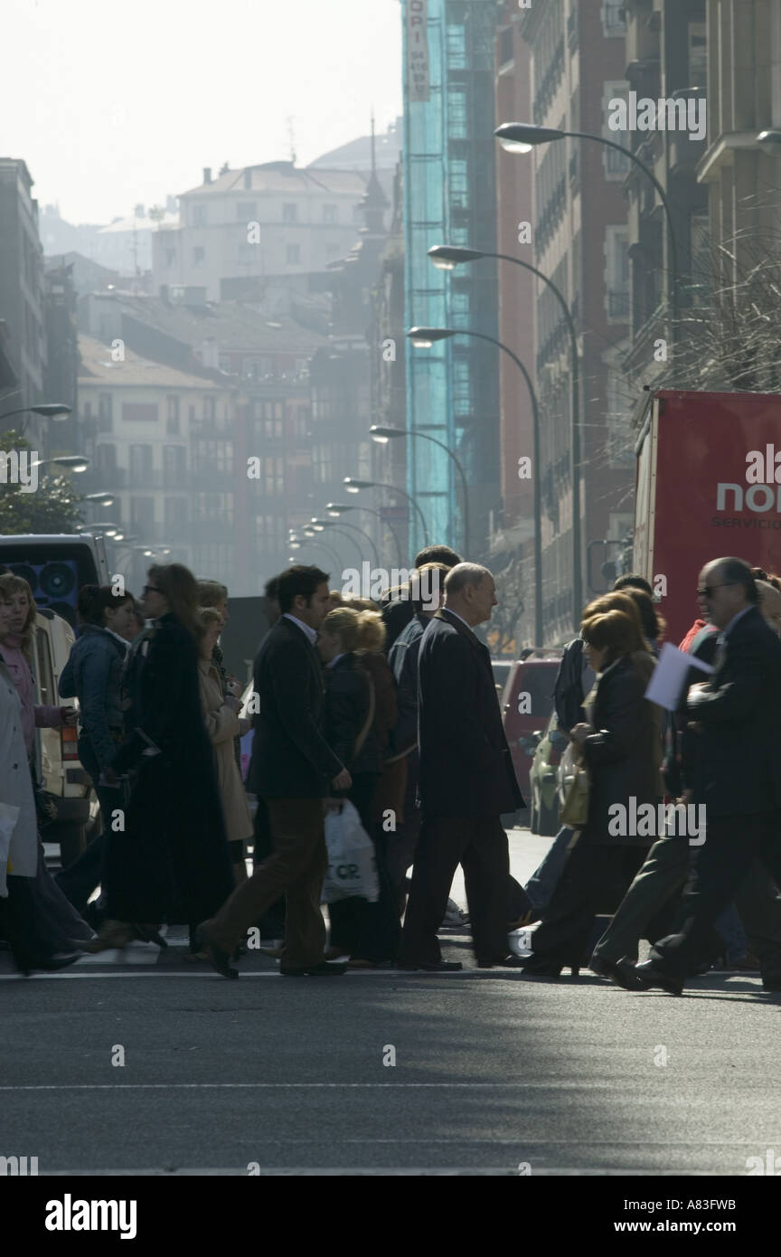 Crowd of Spanish people backlit by the morning sun cross a busy street ...