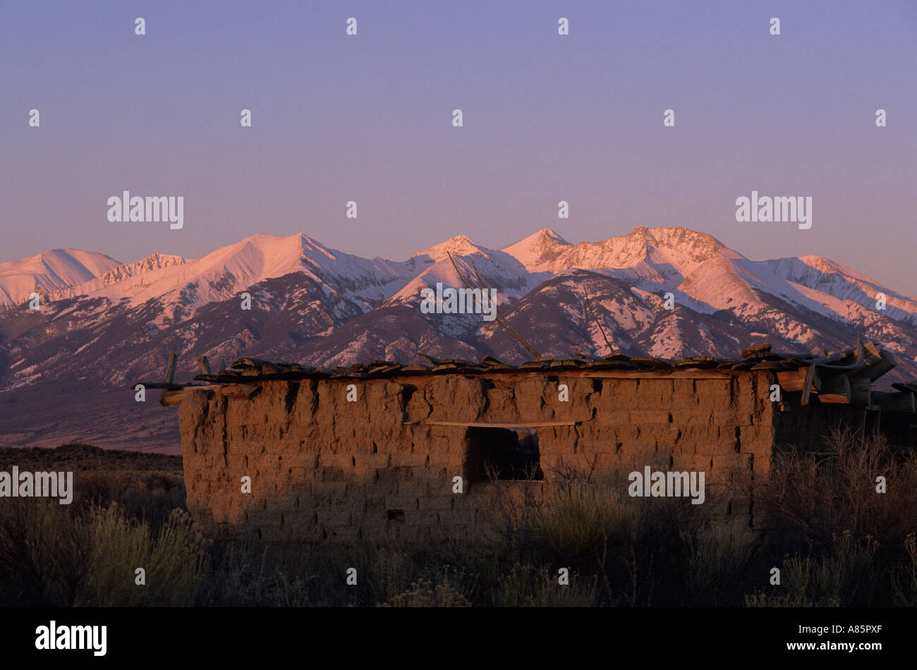 A mud brick cabin nestled in a valley below a snowcapped mountain range ...