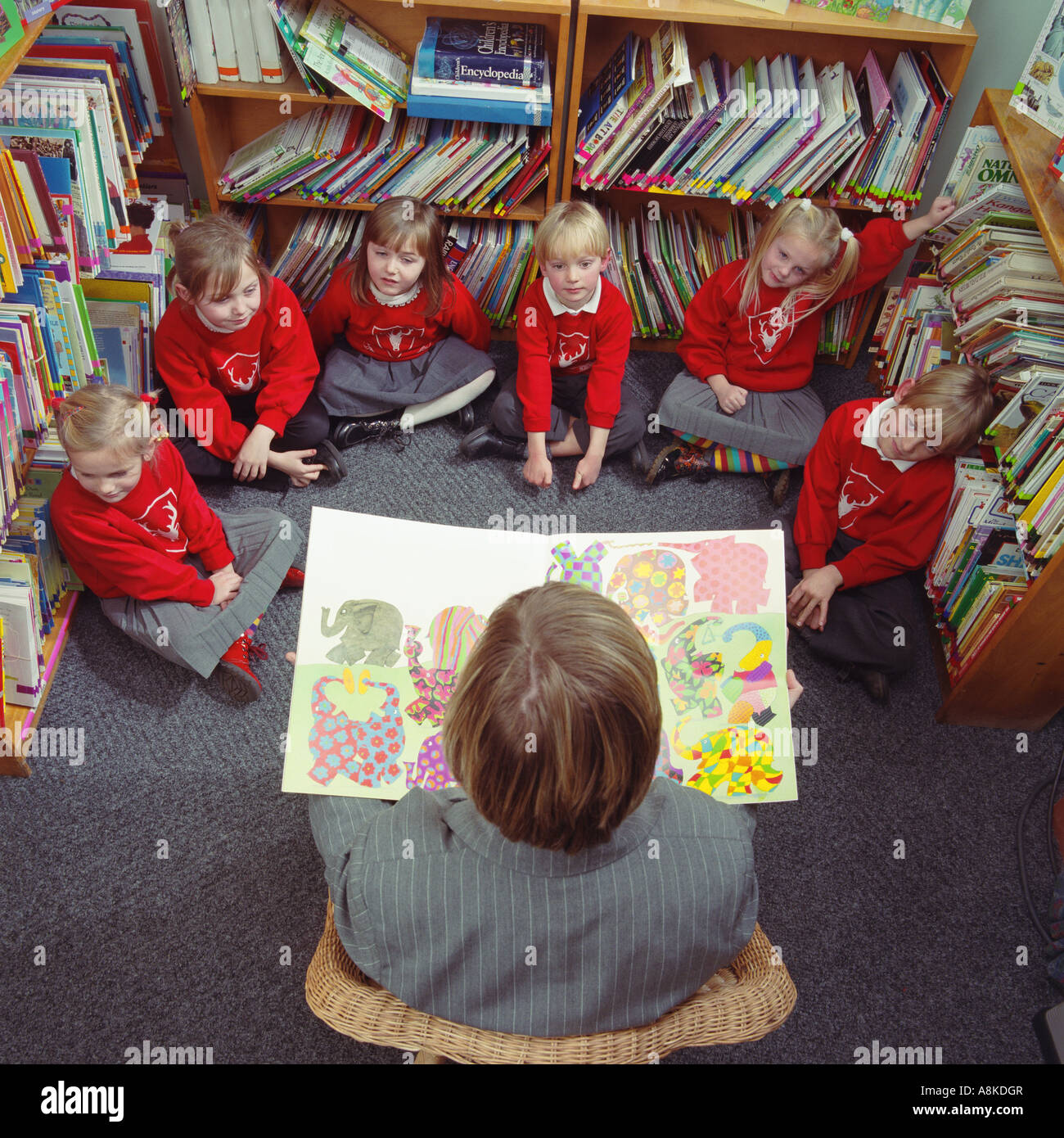 A teacher reading a book to a group of children child with arm raised ...