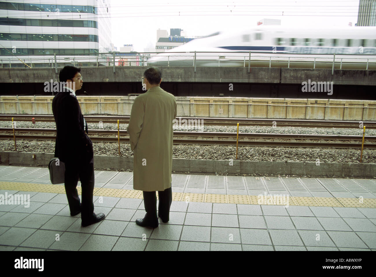Business men wait and talk on a platform as a JR700 series shinkansen (bullet train ) streaks ...