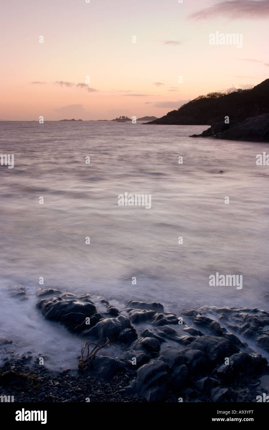 Misty waves crash upon a rocky pebble beach at sunset creating eery ...