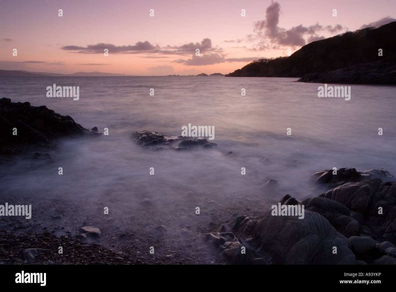 Misty waves crash upon a rocky pebble beach at sunset creating eery ...
