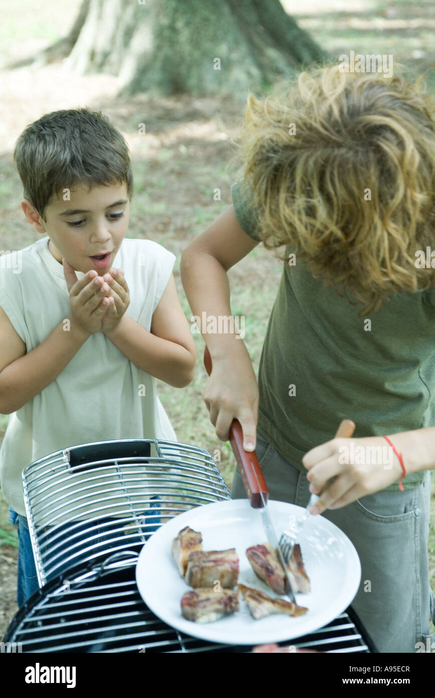 Boy cutting up piece of grilled meat while second boy watches Stock ...