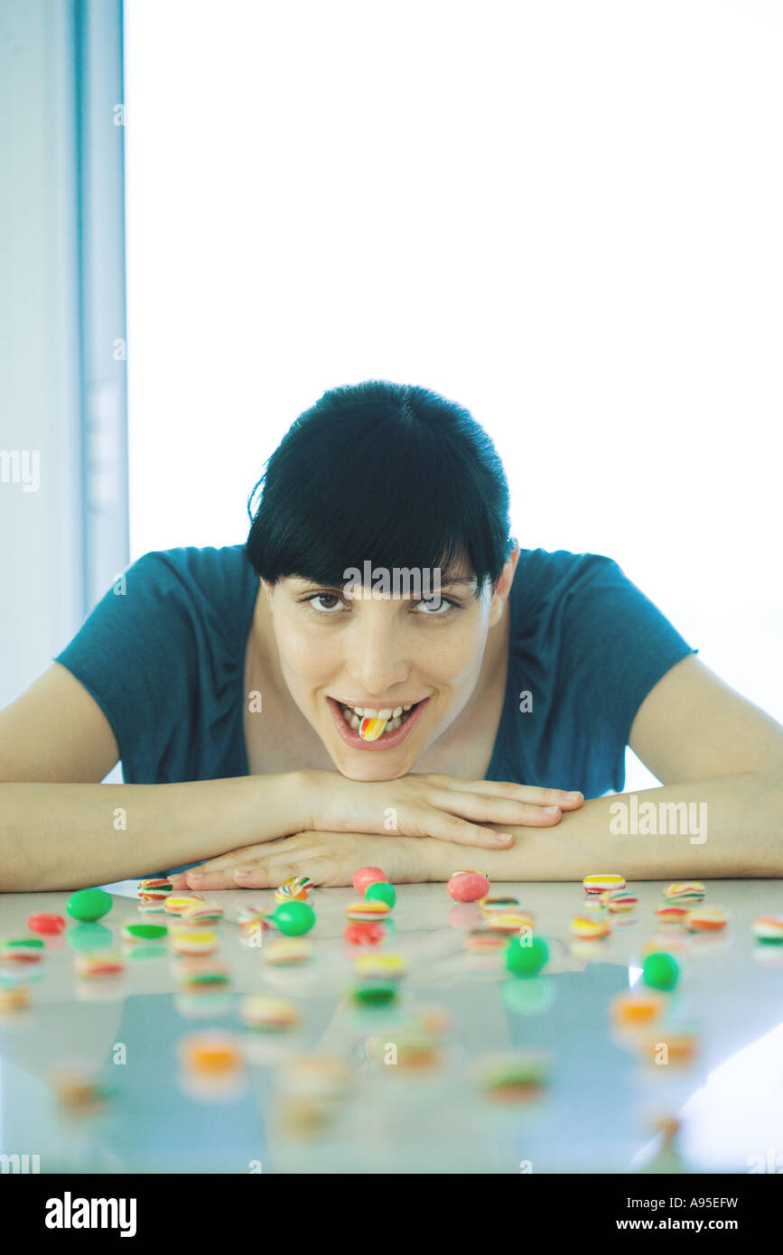 Young woman sitting at table covered with candy, holding piece of candy ...
