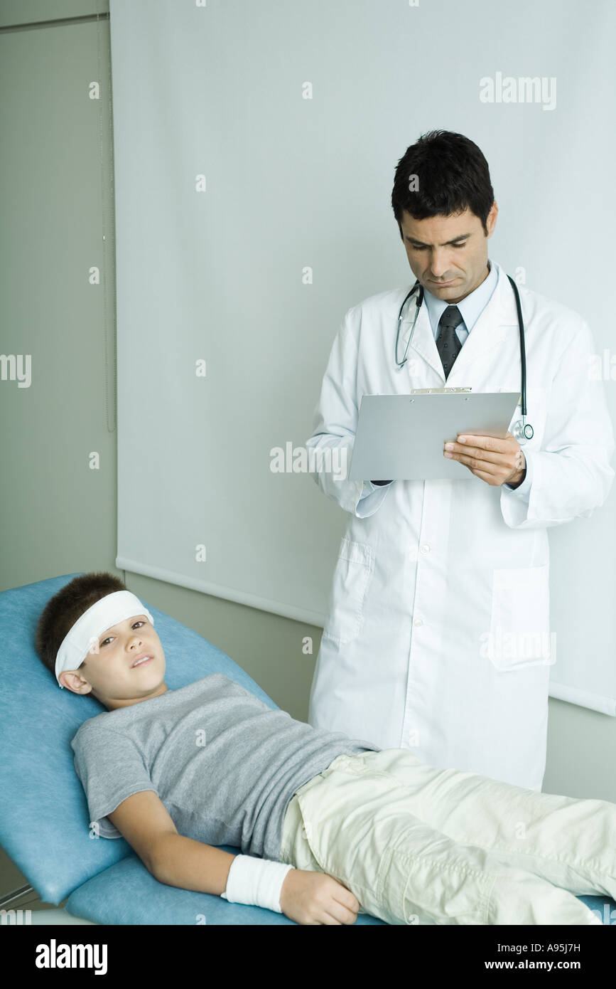 Boy lying on examination table with bandage on forehead and arm, doctor ...